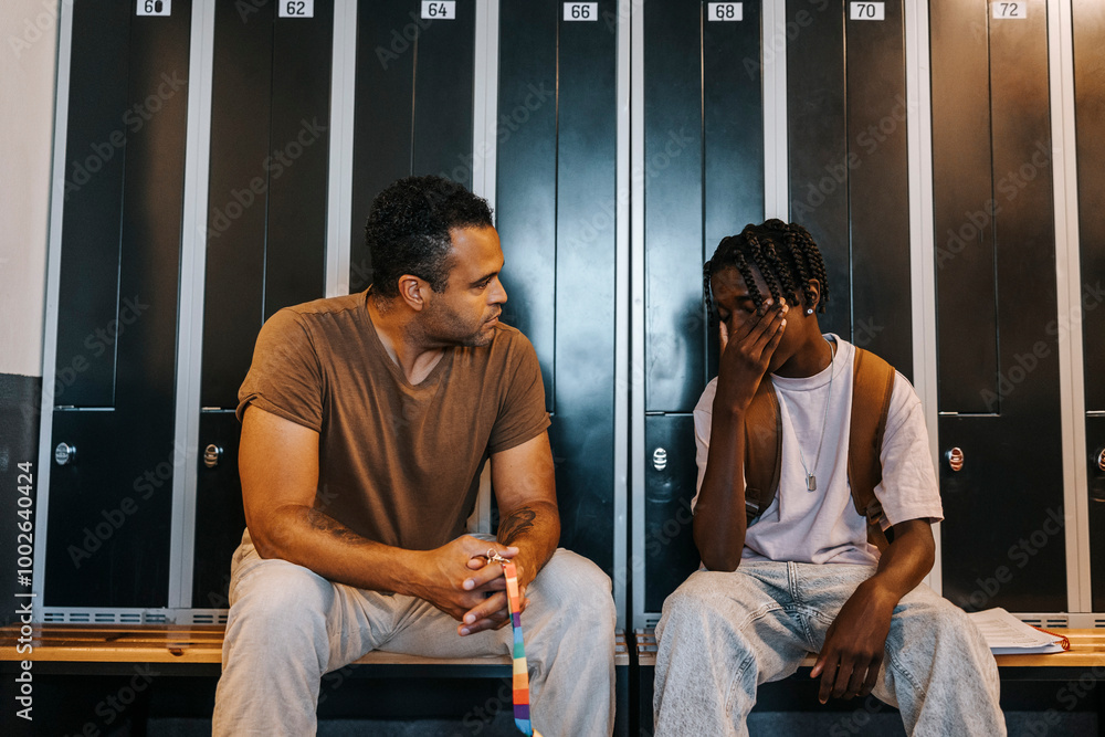 Male sports coach consoling sad teenage boy sitting on bench in locker ...