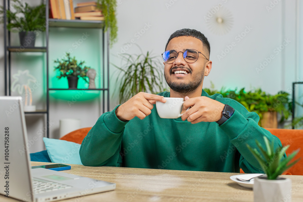 Handsome smiling Indian man working on laptop computer drinking a cup ...