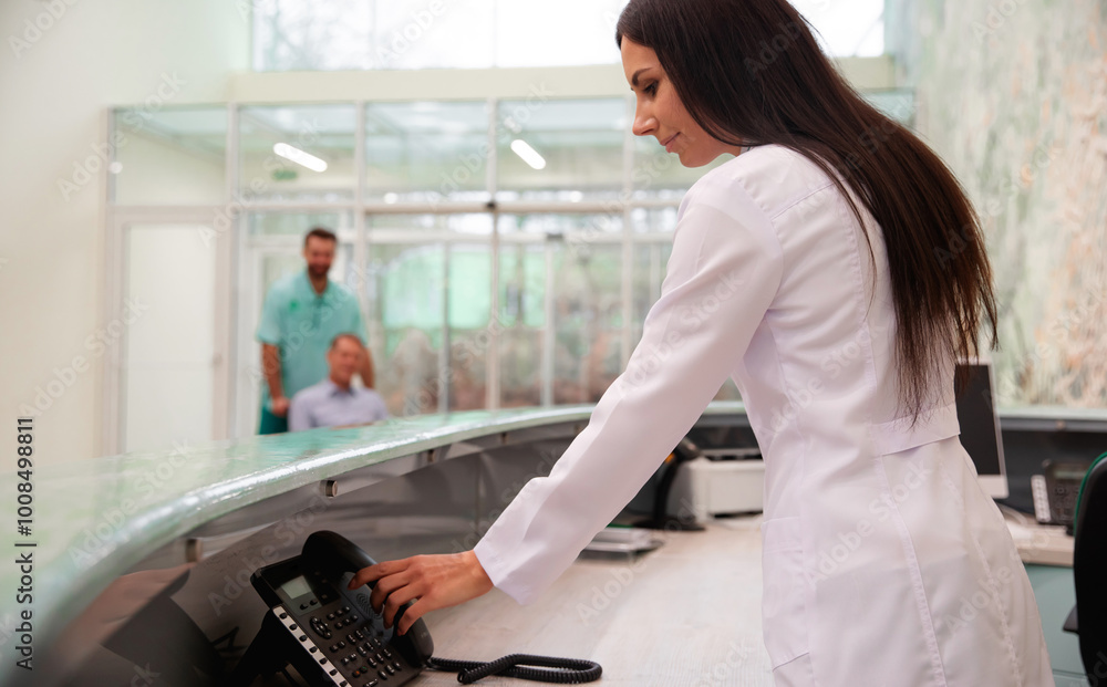 A female medical receptionist in a white coat answers the phone at a ...