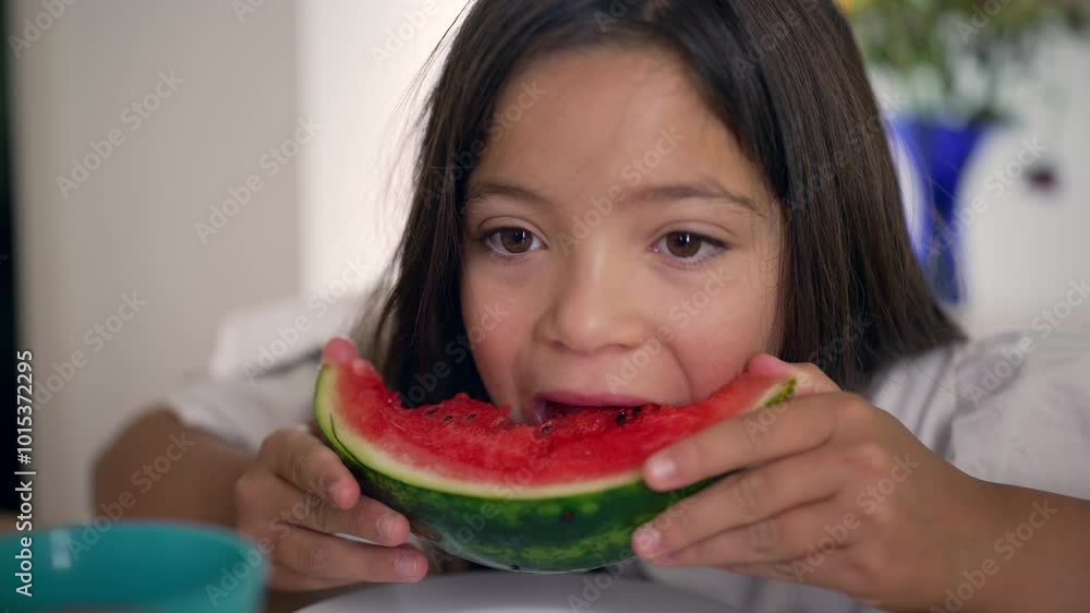 Girl biting into a slice of watermelon at a dining table, showing ...