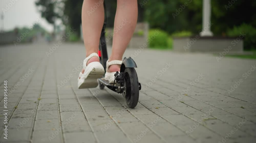 Close-up of a woman riding a black scooter on a paved walkway, her legs ...