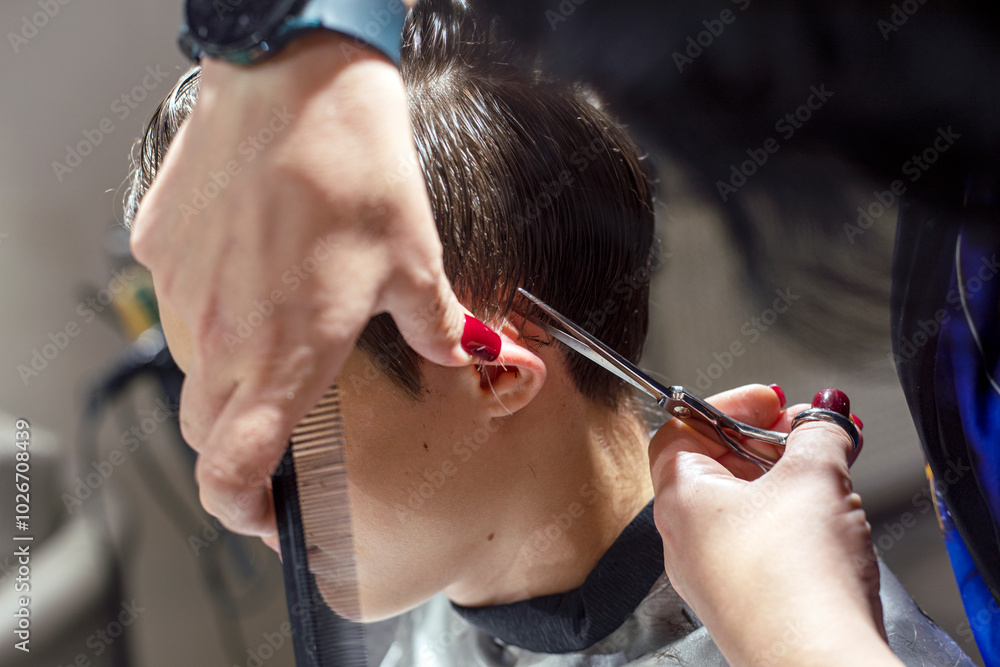 barber cuts the hair near the ears of a teenager with scissors in a ...