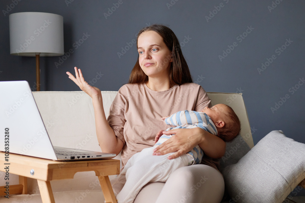 Puzzled woman sitting on couch with her baby in hands talking on video ...
