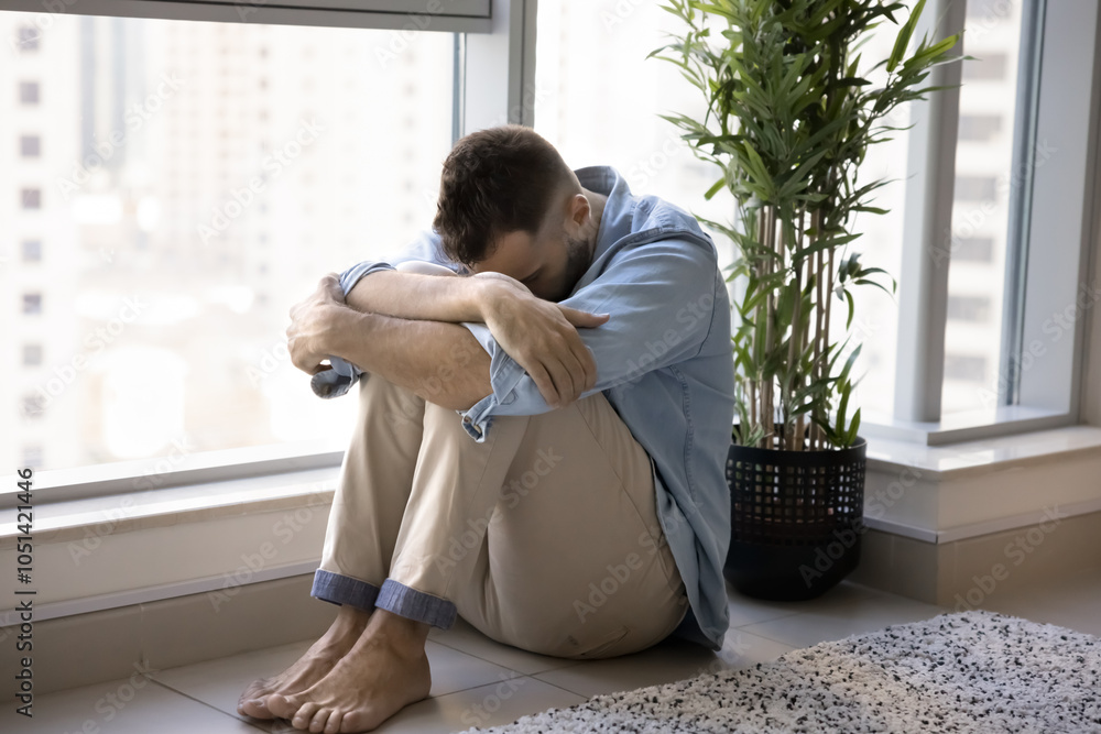Depressed unhappy young man sitting at window on floor, bending head ...