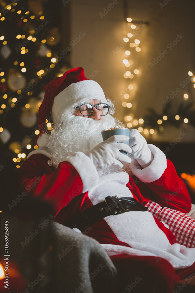 Jolly and relaxed Santa Claus with eyeglasses sits by fireplace, drinks ...