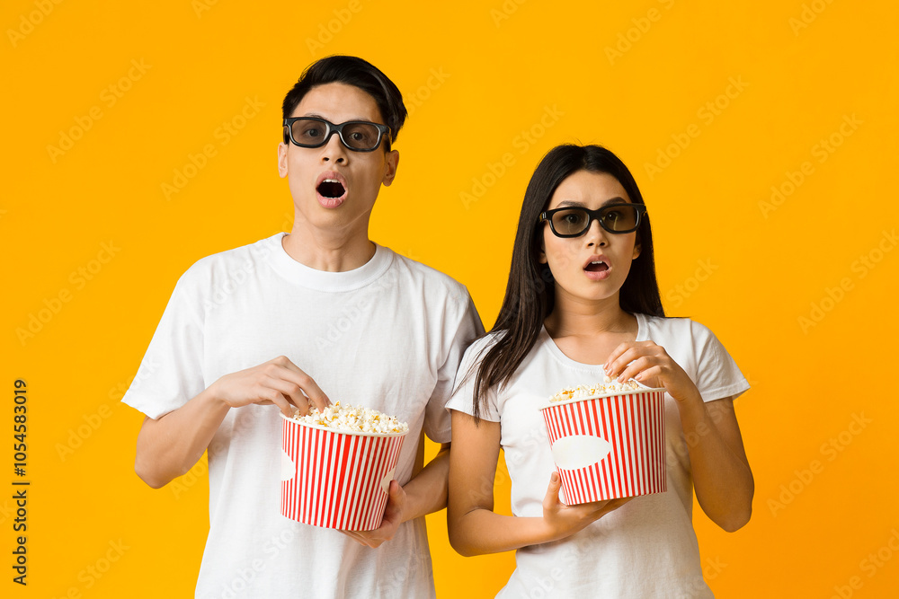 Young stunned asian couple in 3d glasses watching scary movie, holding bucket of popcorn, yellow ...