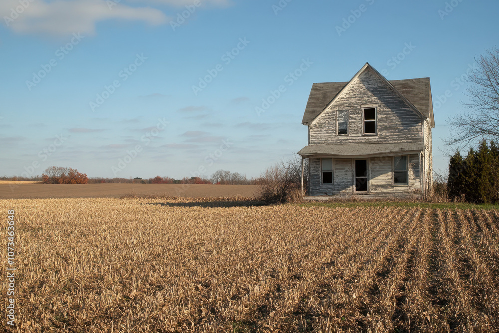 An abandoned farmhouse stands in an empty field under a clear blue sky ...