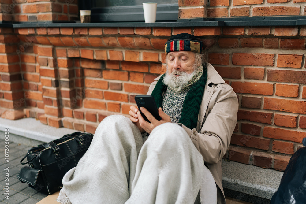 Relaxed aged homeless man with beard using smartphone sitting sidewalk ...