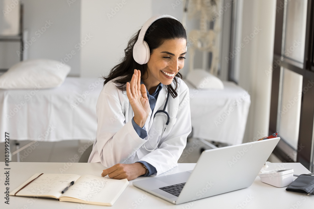 Smiling young 30s female doctor start video call using application, sit at desk look at laptop ...