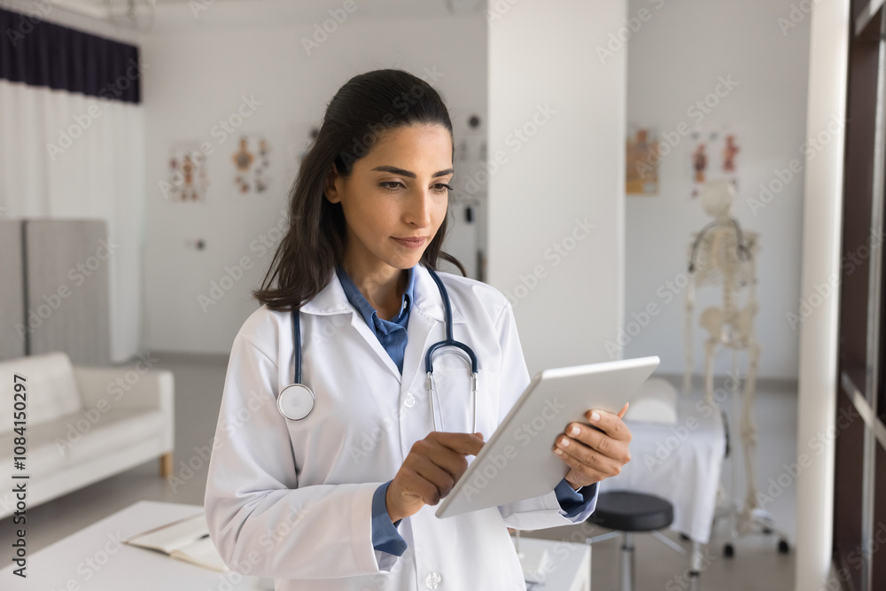 Focused young Latina female doctor wear uniform using tablet standing ...
