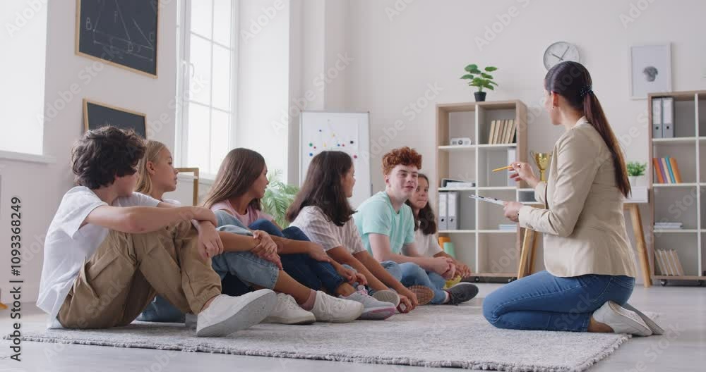 Smiling female teacher sitting on carpet in classroom, leading lesson ...