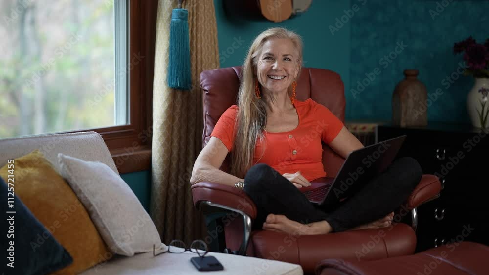 Mature woman with long blonde hair, wearing an orange shirt, sits in a ...