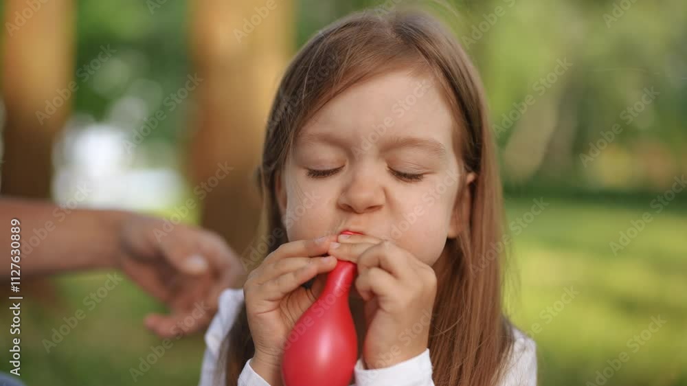 A young girl joyfully blowing into a bright red balloon, embodying ...