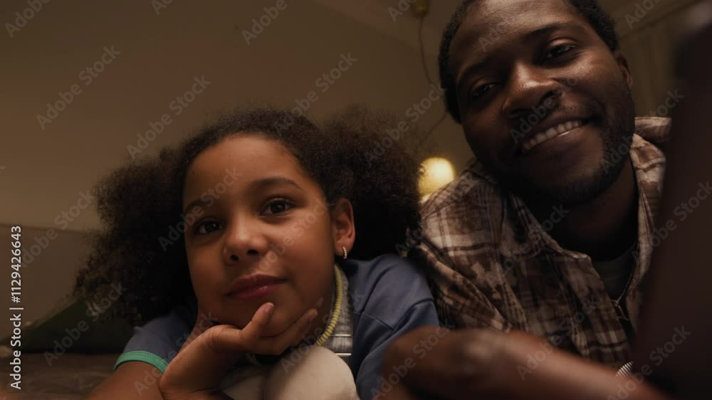 POV shot of little biracial girl lying in bed with his happy young Black dad wearing comfy home ...