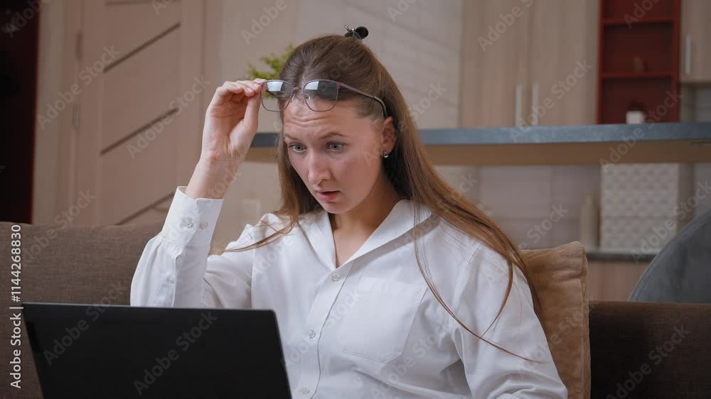 Young woman adjusting her glasses while frowning at laptop screen ...