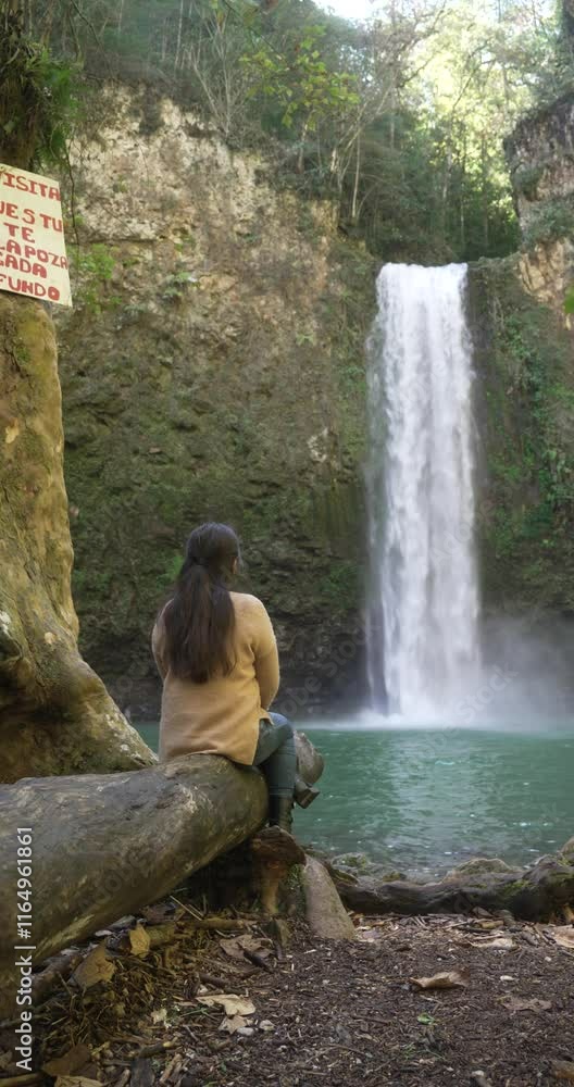 Mexican young girl sitting and watching a waterfall. Theres a sign in ...